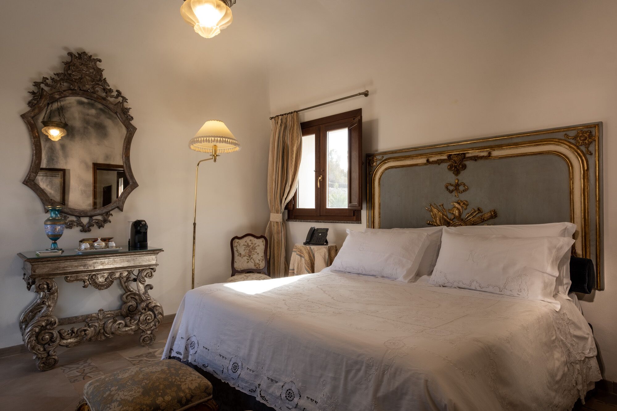 Quarto com uma cama grande, cabeceira elaborada, um espelho, um abajur e uma pequena mesa com luminária.