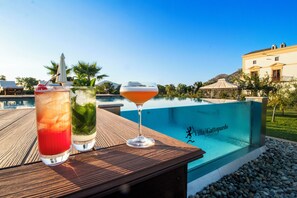Mesa à beira da piscina com três bebidas, uma piscina e uma villa chamada 'Villa Gattopardo' ao fundo.
