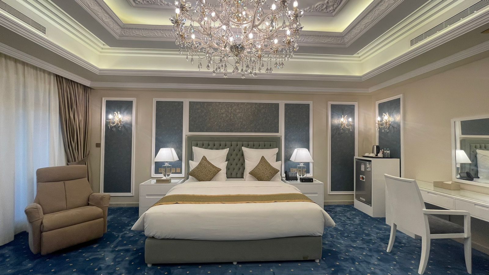 Quarto de hotel com uma cama grande, uma escrivaninha, uma cadeira, um candelabro e uma janela ampla.