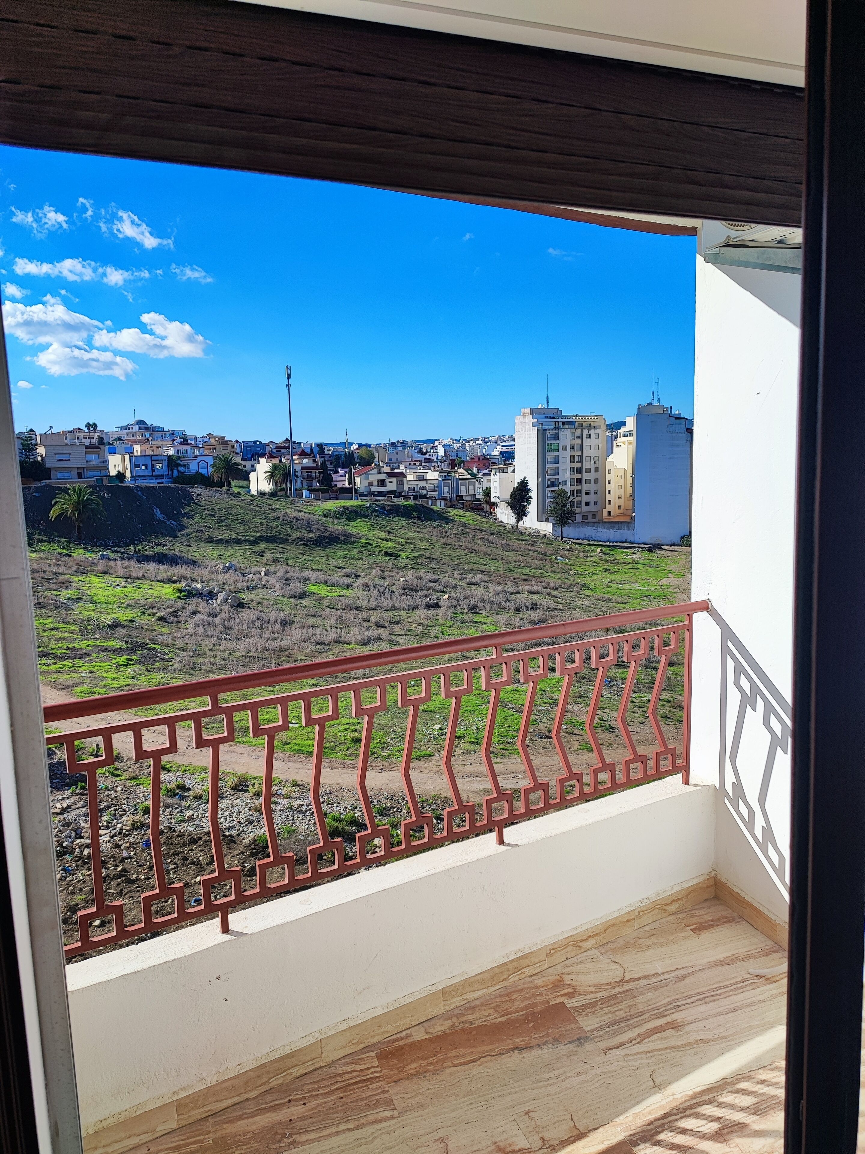 Varanda com guarda-corpo de metal vermelho, vista para o horizonte urbano com edifícios e céu azul.