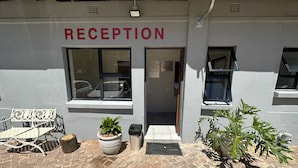 Uma área de recepção com uma placa indicando "RECEPTION" acima da porta, um banco, plantas em vasos e uma janela com vista para carros estacionados.