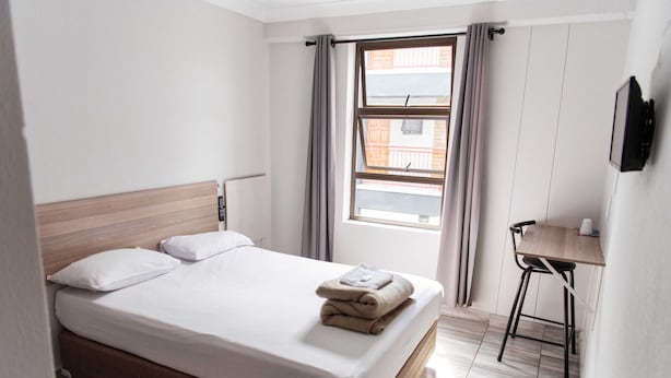 Uma cama bem arrumada com cabeceira de madeira, uma pequena mesa com uma cadeira e uma janela com cortinas.