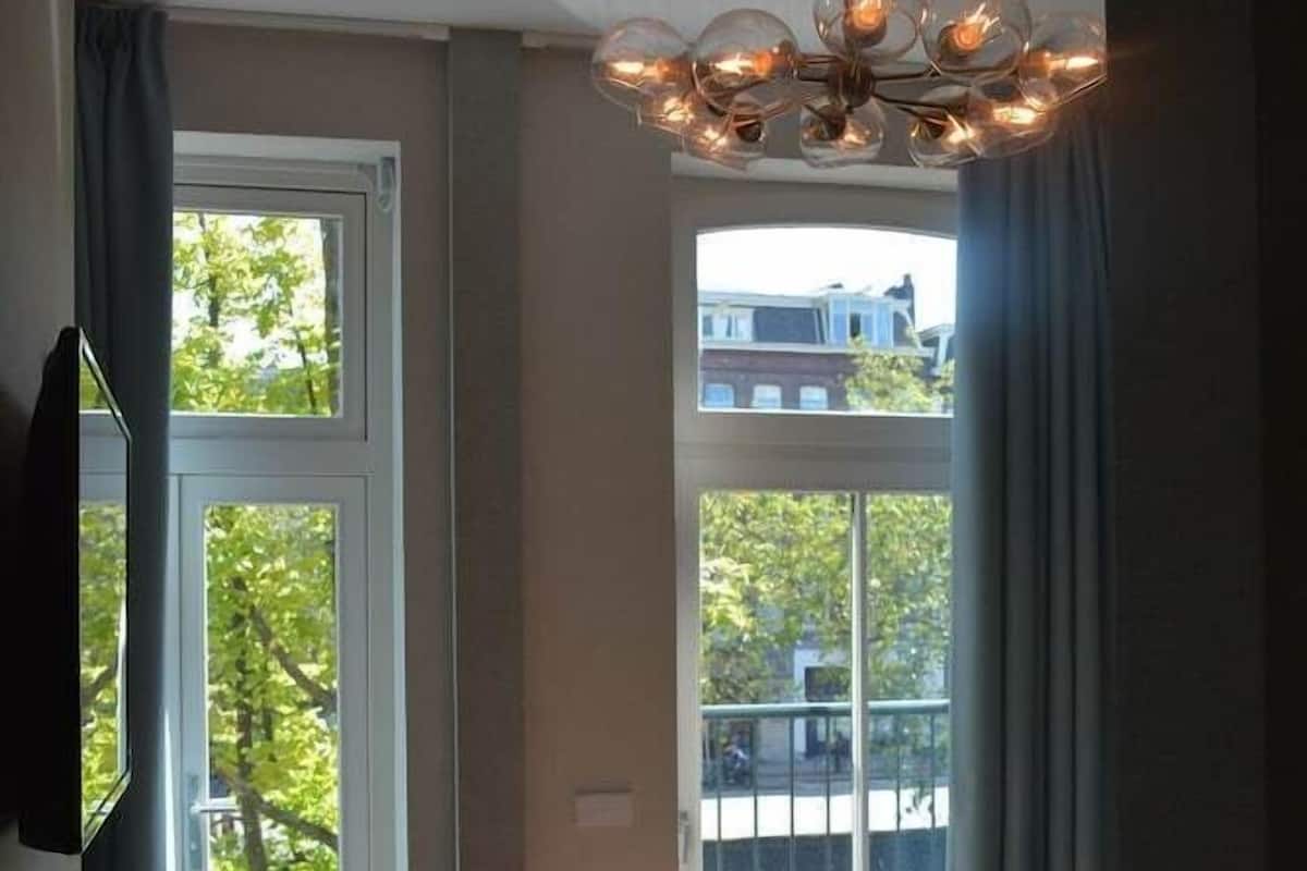 Quarto com cama, cadeira, luminária pendente e janelas amplas.