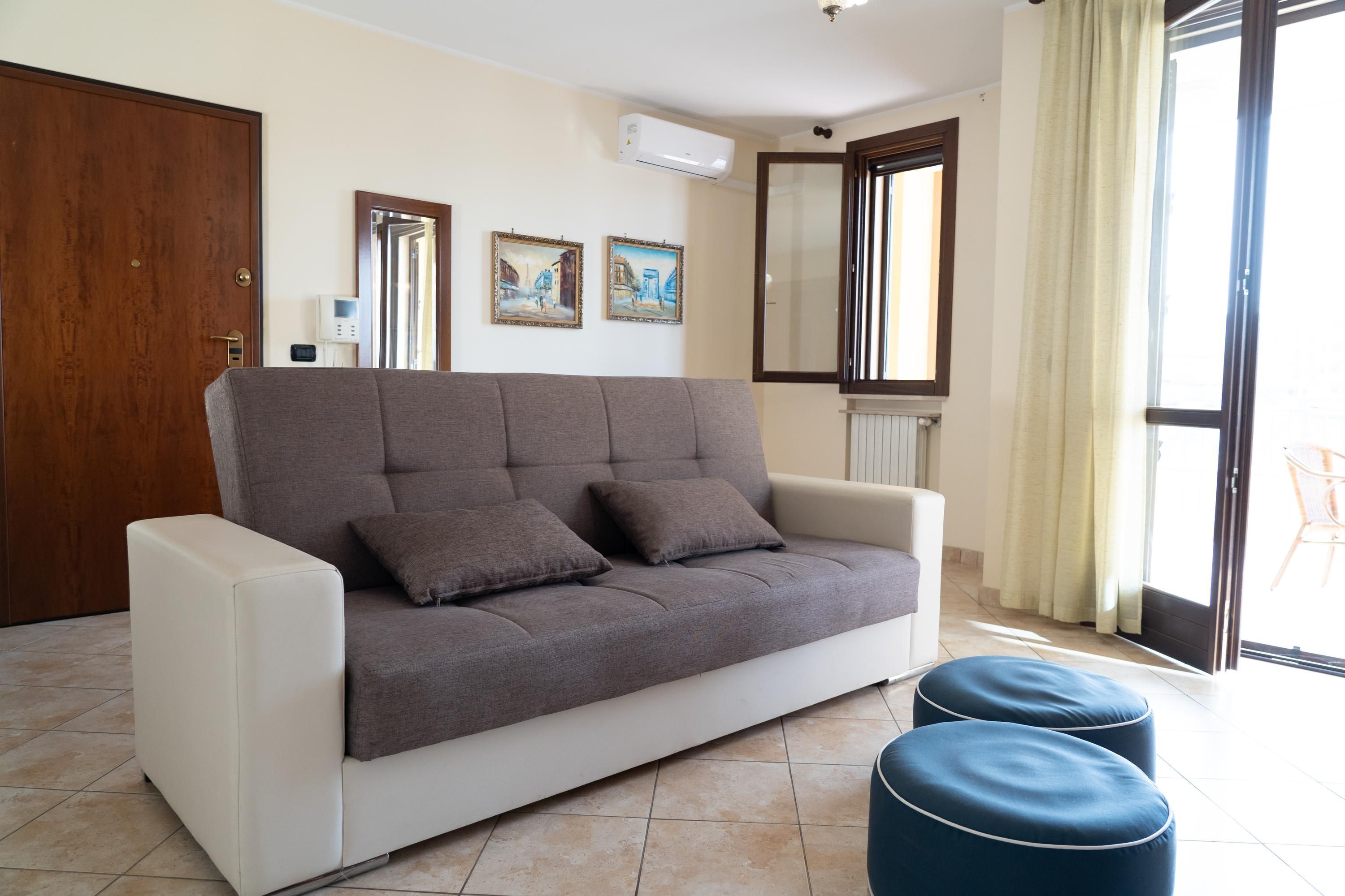 Uma sala de estar com um sofá cinza, dois pufes azuis, uma porta de madeira e uma janela com cortina.