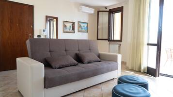 Uma sala de estar com um sofá cinza, dois pufes azuis, uma porta de madeira e uma janela com cortina.