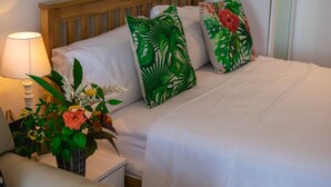 Cama bem arrumada com lençóis brancos e duas almofadas decorativas com estampas de folhas tropicais.