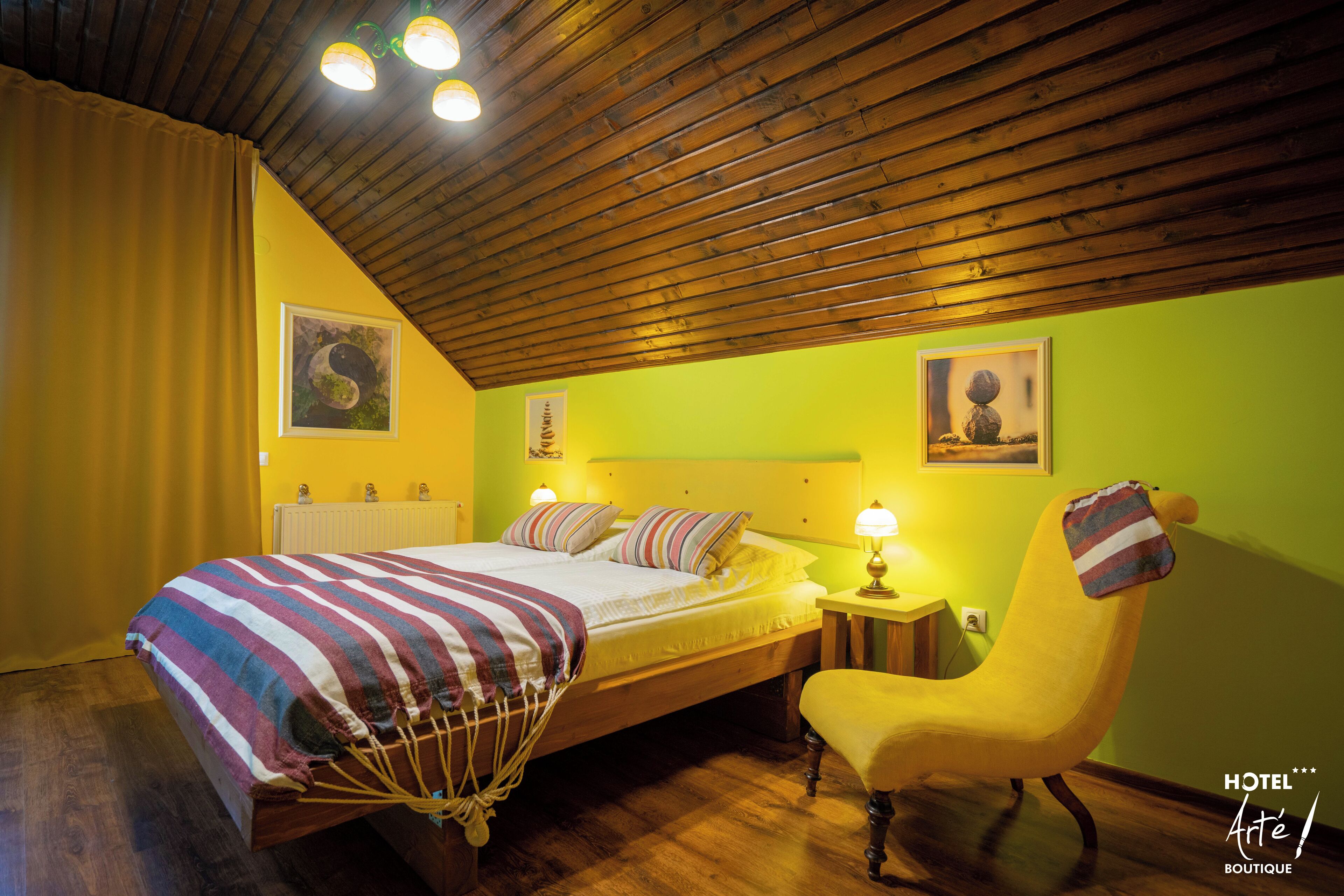 Quarto de hotel com teto de madeira, uma cama com roupa de cama listrada, uma poltrona amarela e duas luminárias fixadas na parede.