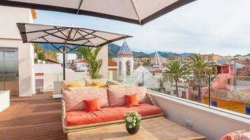 Um terraço no telhado com piso de madeira, um sofá com almofadas estampadas, uma mesa com um vaso de flores e vista para uma paisagem urbana com edifícios e uma ponte.