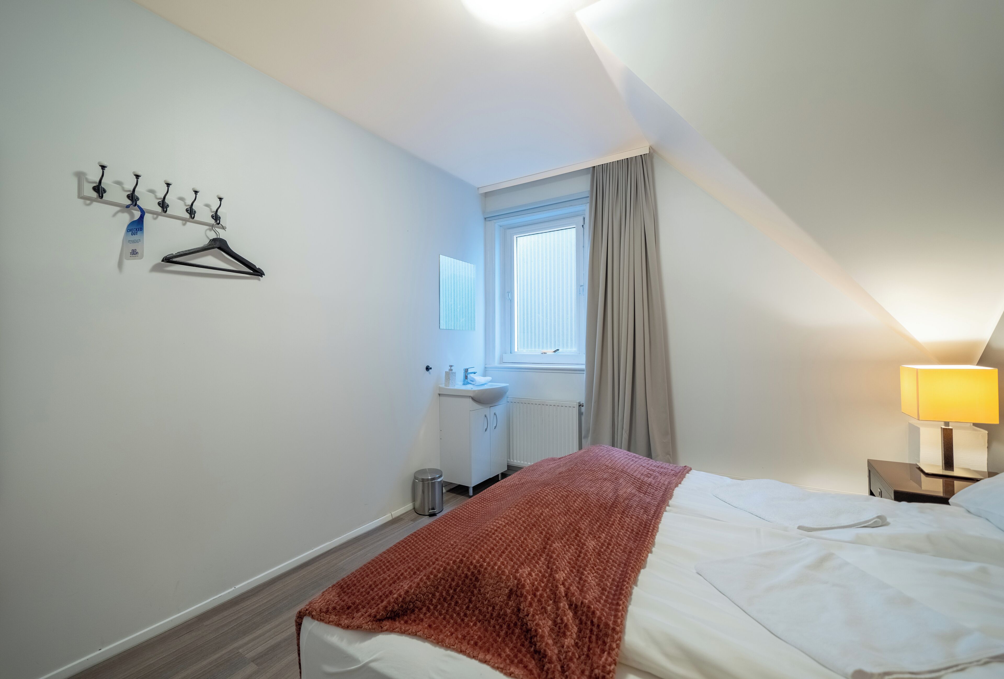 Quarto de hotel com cama, janela com cortinas, mesa de cabeceira com abajur, cabideiro com cabides e um pequeno armário com pia.