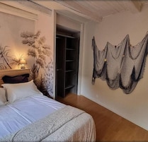 Quarto com cama, mesa de cabeceira, luminária, prateleira na parede e uma janela grande.