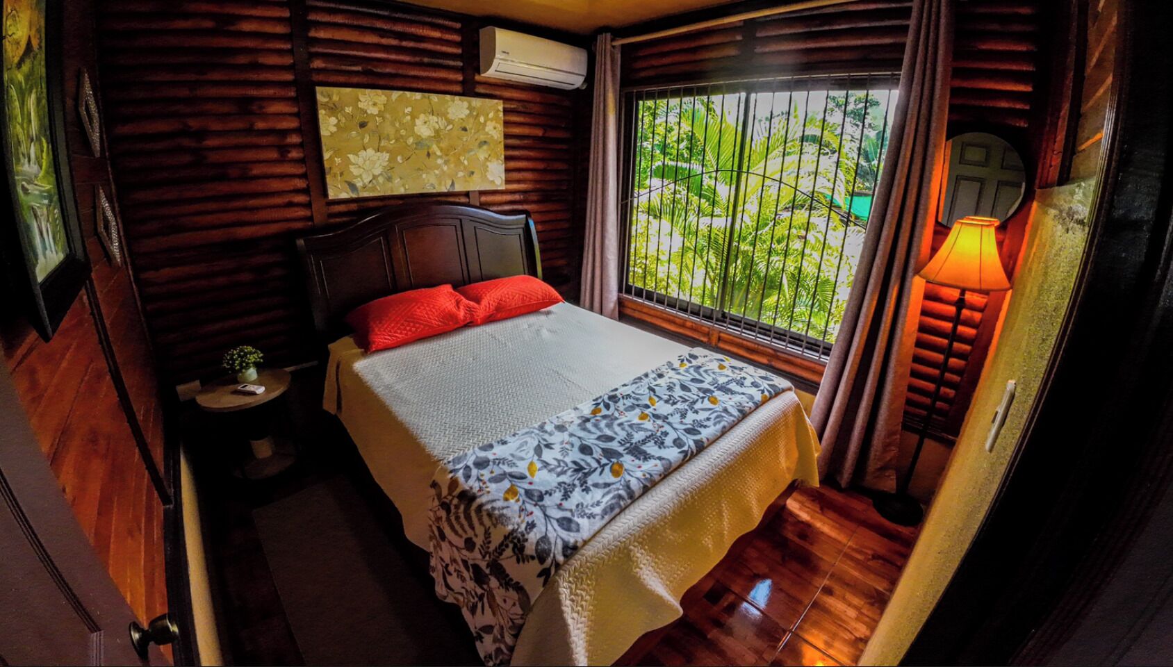 Quarto com cama, mesa de cabeceira, janela com vista para o verde e aparelho de ar condicionado.