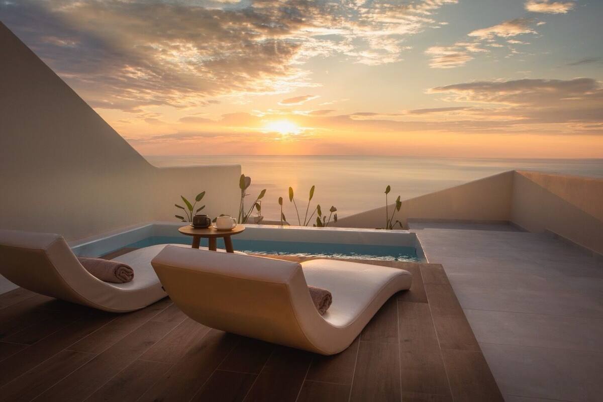 Terraço moderno no telhado com duas cadeiras de estar, uma mesa pequena e uma piscina, com vista para o pôr do sol sobre o mar.