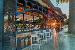 Uma área de bar ao ar livre com banquetas brancas, uma estrutura com telhado de palha e vista para o pôr do sol.