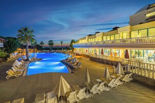 Uma área de piscina com espreguiçadeiras e guarda-sóis, um edifício com varanda e céu claro.
