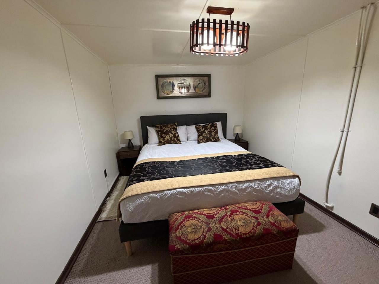 Quarto bem arrumado com cama, mesas de cabeceira, um banquinho e um lustre.