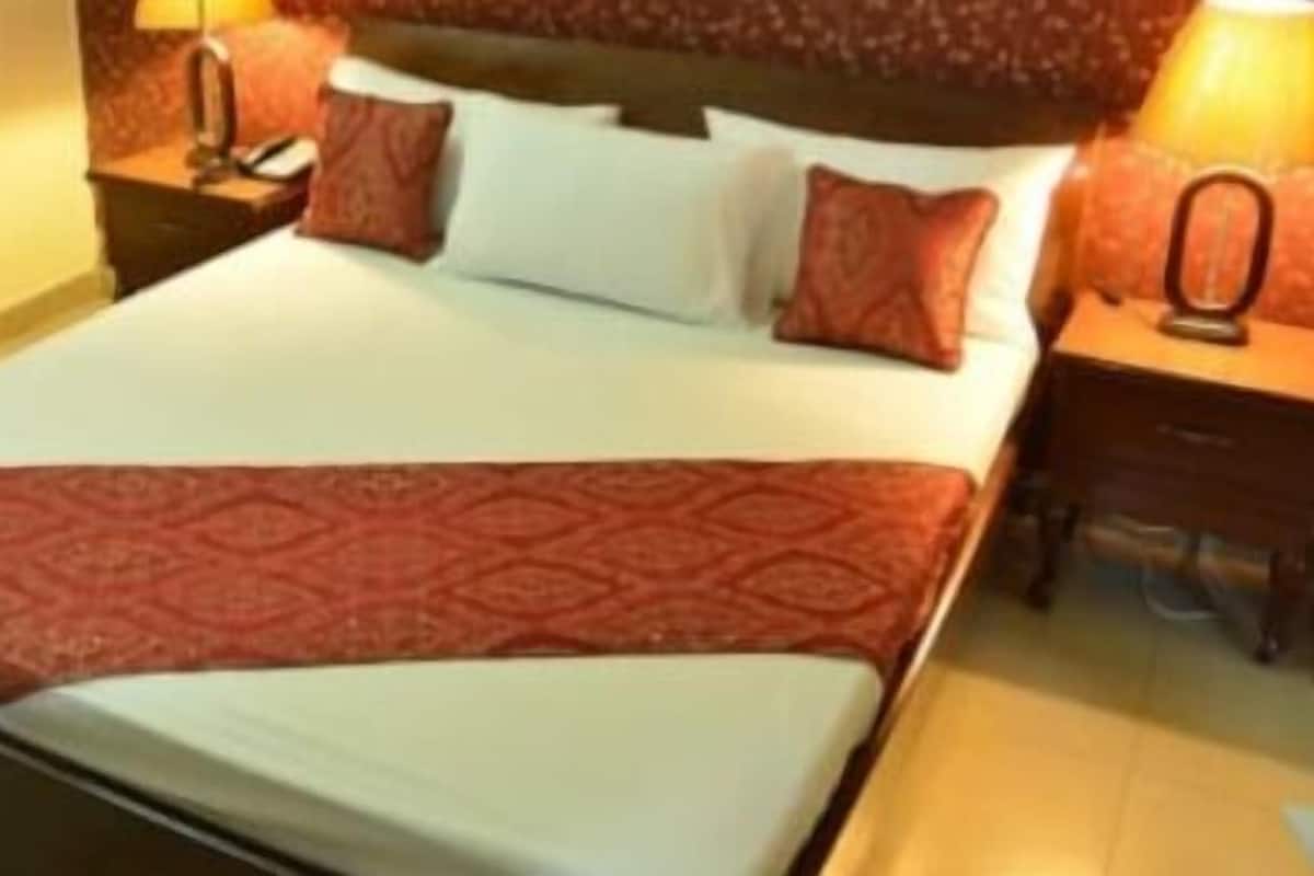 Uma cama bem arrumada com colcha estampada, duas mesas de cabeceira com abajures e uma cabeceira com padrão decorativo.