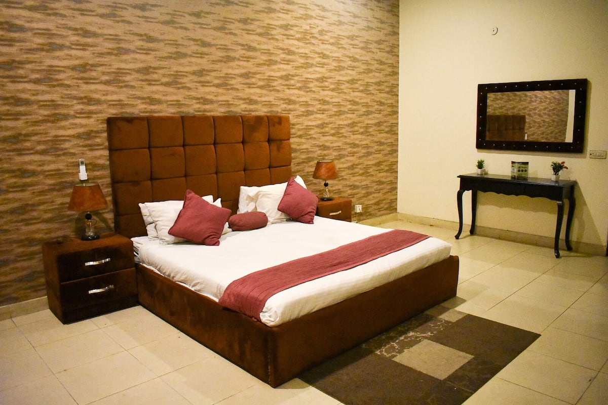 Uma cama bem arrumada com cabeceira marrom, duas mesas de cabeceira com luminárias, uma mesinha com objetos decorativos e um espelho na parede.