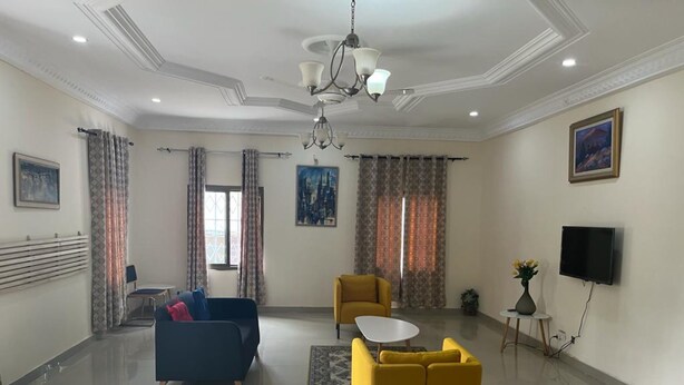 Sala de estar ampla com um sofá azul, uma poltrona amarela e uma mesinha de centro. Há uma televisão fixada na parede e um quadro acima.