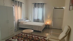 Quarto com cama, cadeira, mesinha, guarda-roupa e janela com cortinas.