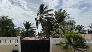 Uma entrada fechada com um caminho de concreto que leva a um edifício, cercado por palmeiras e um muro branco.