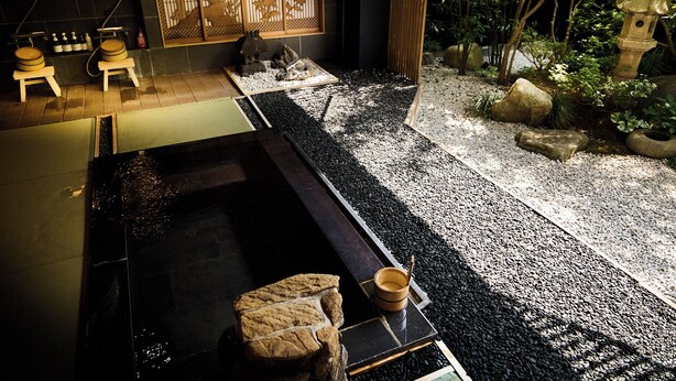 Um interior em estilo japonês tradicional, com tatames, uma mesinha baixa e uma pequena área de jardim com pedras e um espelho d'água.
