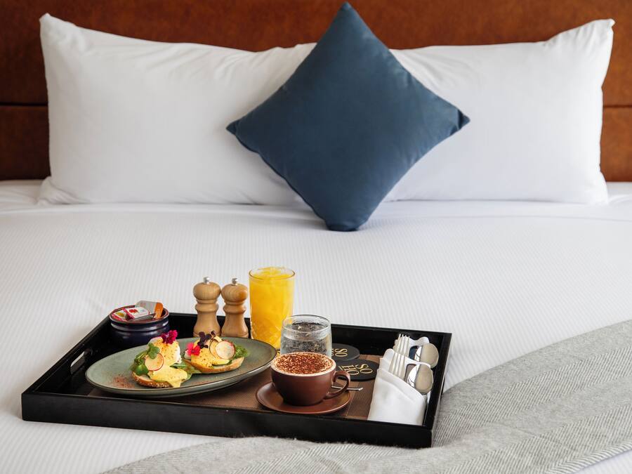 Uma bandeja de café da manhã bem arrumada com ovos, torradas e suco em uma cama com almofadas brancas e azuis.