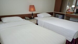 Quarto de hotel com duas camas de solteiro, uma mesa de cabeceira com abajur, telefone e uma prateleira pequena com objetos.