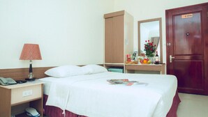 Quarto de hotel com cama, mesa de cabeceira com abajur, guarda-roupa, espelho, porta e uma pequena mesa com cesta de frutas e uma garrafa.