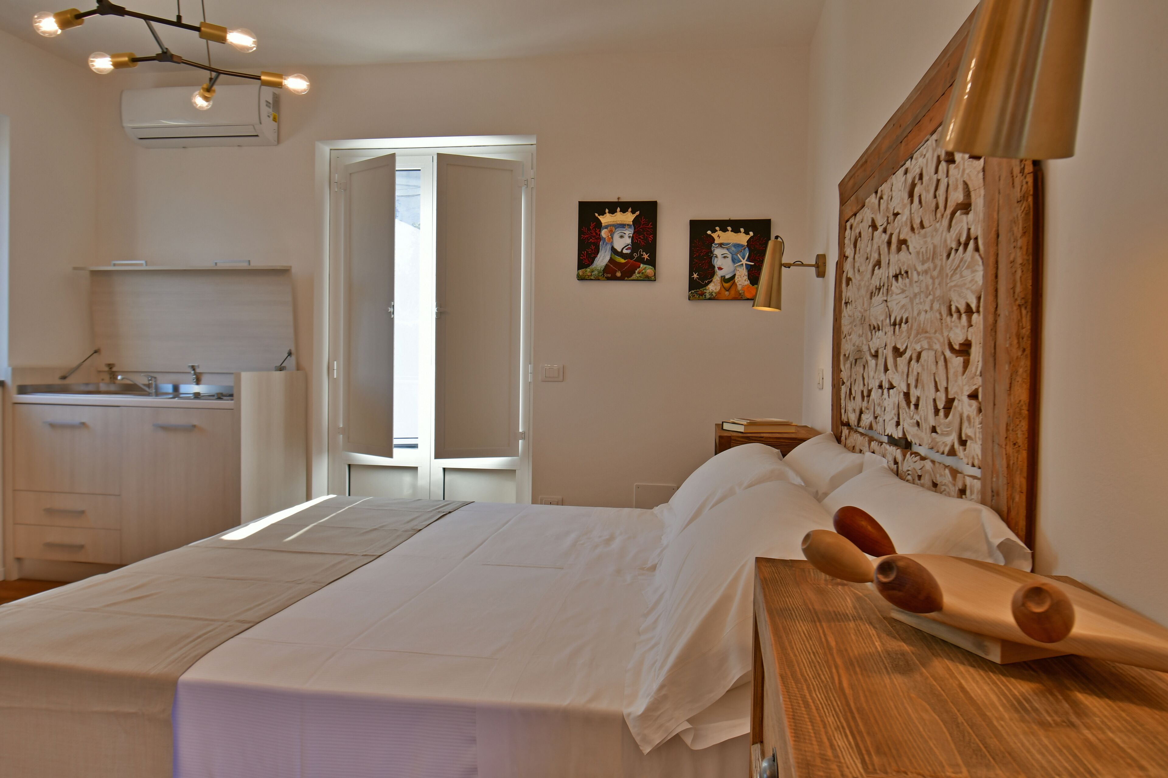 Quarto com cama, cabeceira de madeira e mesa de cabeceira com abajur. Há uma porta que dá para um banheiro e uma cozinha.