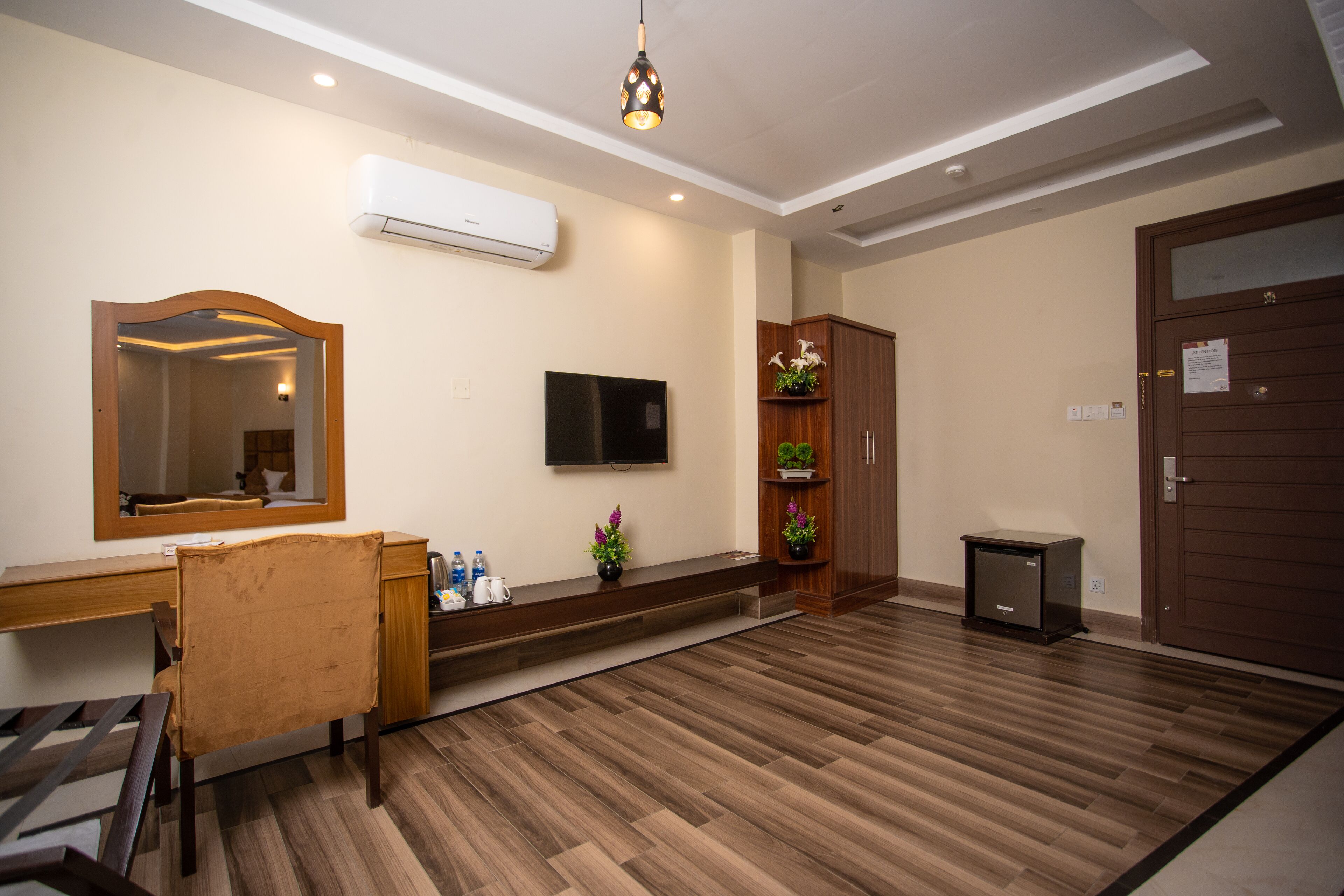 Quarto de hotel com piso de madeira, um espelho grande, televisão de tela plana, um pequeno armário com plantas e uma porta com tranca.