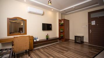 Quarto de hotel com piso de madeira, um espelho grande, televisão de tela plana, um pequeno armário com plantas e uma porta com tranca.