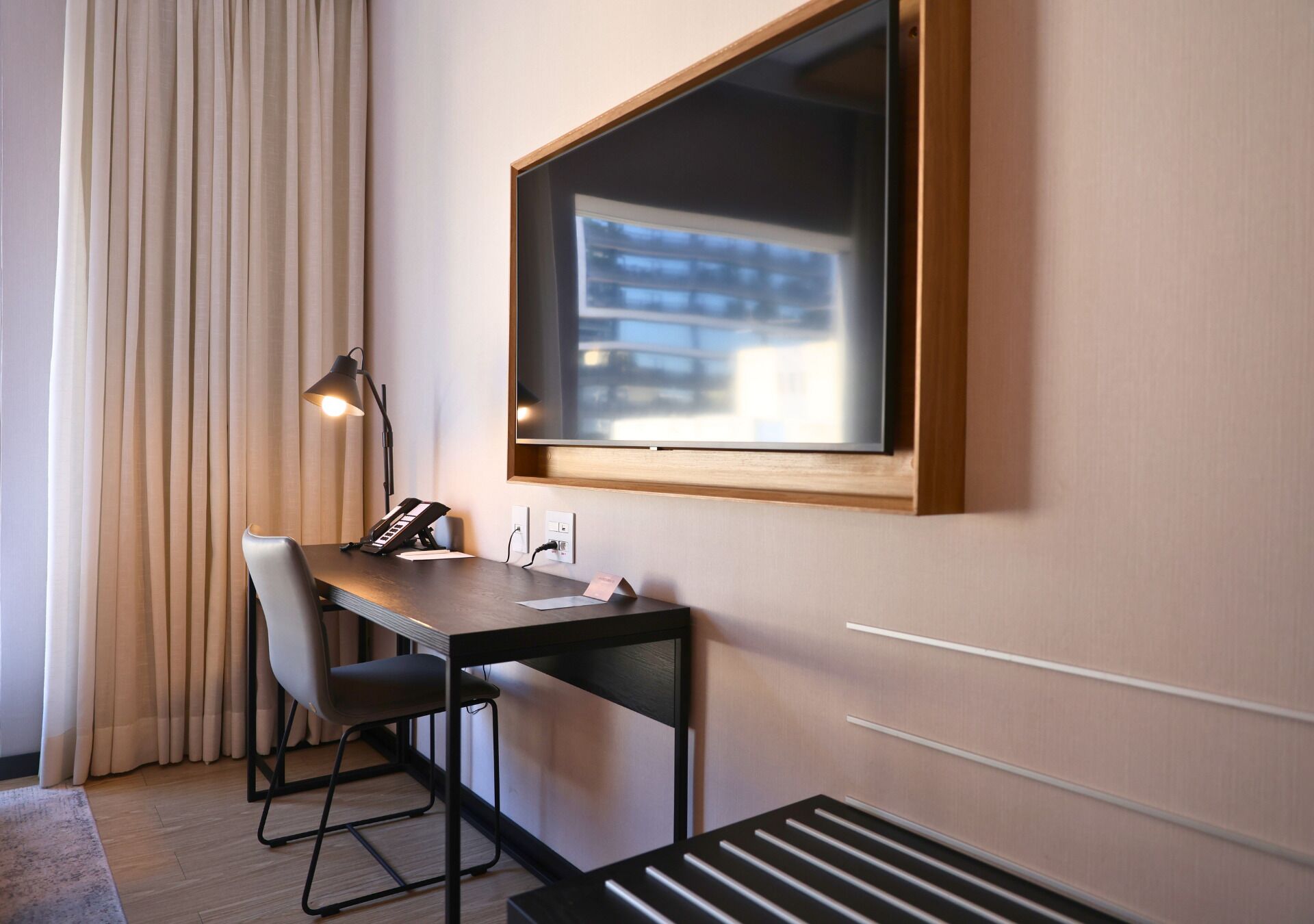 Quarto de hotel com escrivaninha, cadeira, luminária e uma janela grande com vista para edifícios.