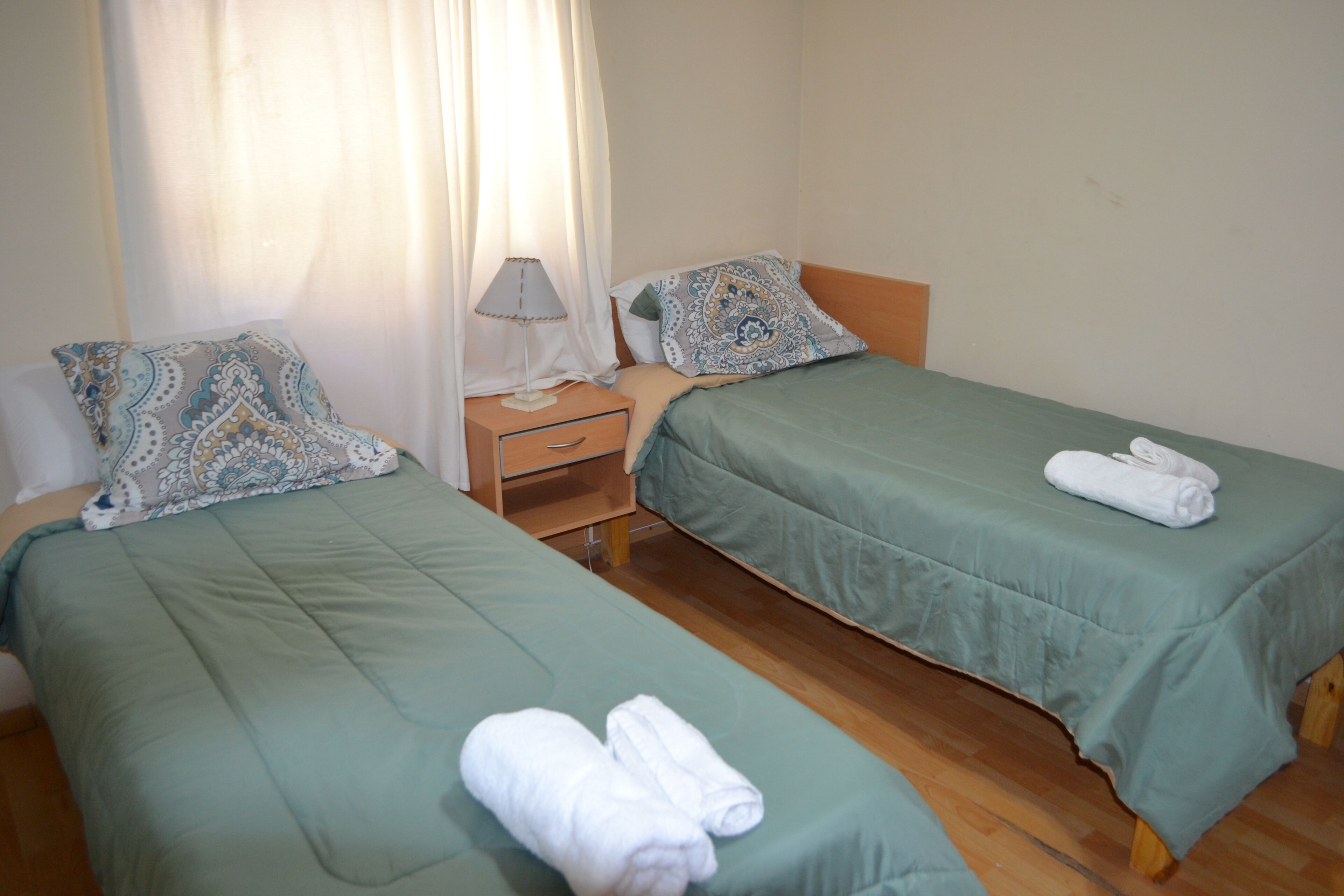 Quarto duplo com duas camas de solteiro, mesa de cabeceira, luminária e janela com cortinas.