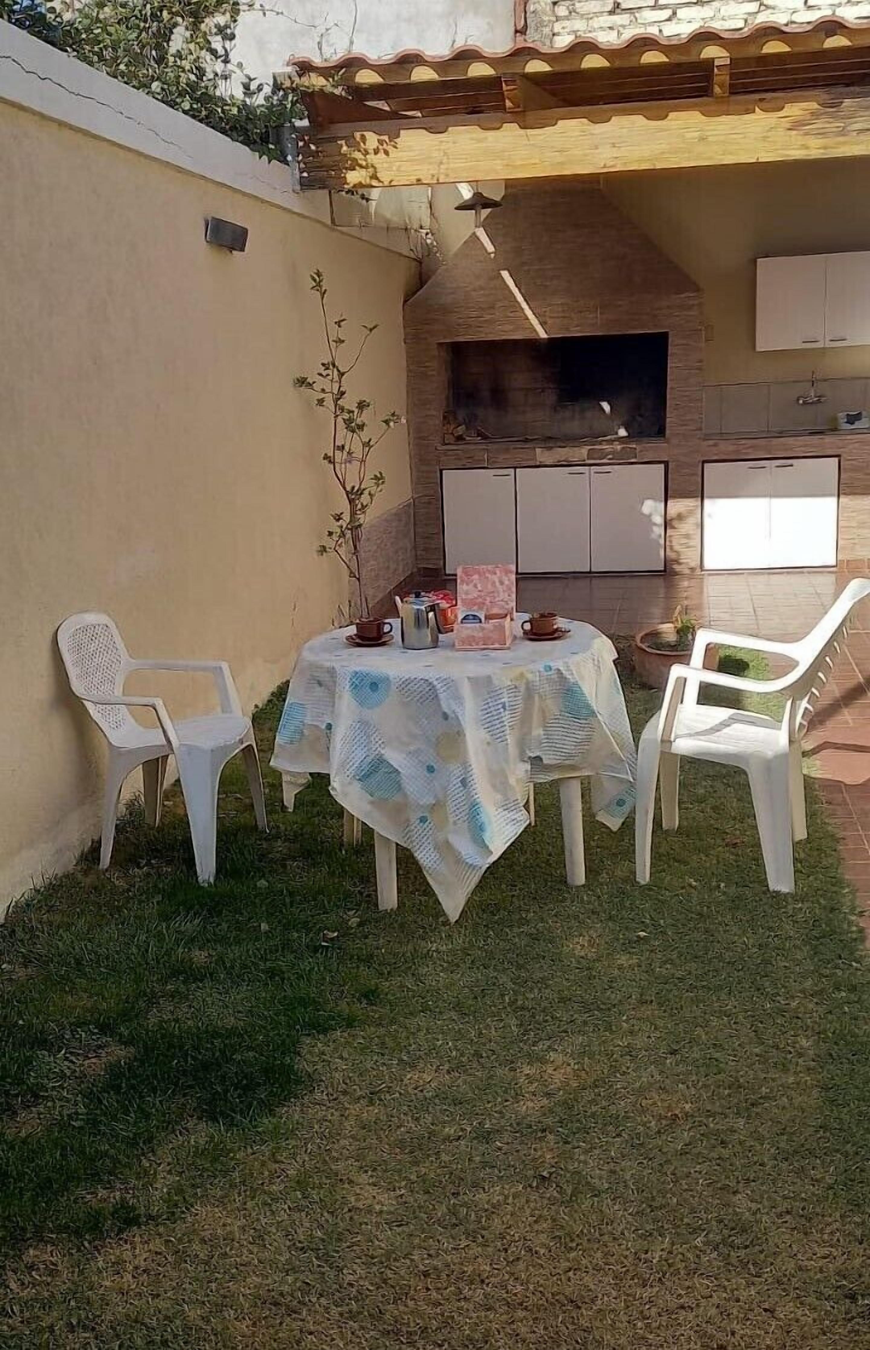 Um pátio com uma mesa posta para dois, rodeado por cadeiras de plástico branco, uma pequena planta e uma área de churrasqueira com armários.