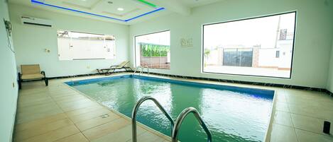 Piscina interior com água azul cristalina, escada de metal e vista para uma área residencial através de amplas janelas.
