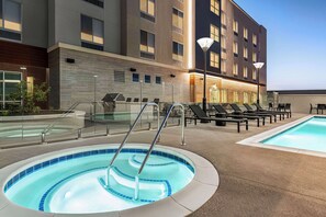 Área de piscina no terraço com espreguiçadeiras e uma banheira de hidromassagem, cercada por um edifício moderno com grandes janelas.