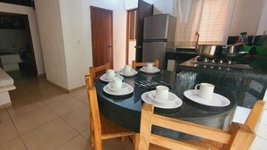 Uma sala de jantar com uma mesa redonda posta para seis, cozinha ao fundo e um pequeno espaço de estar com um banco.
