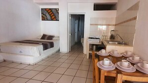Quarto pequeno e limpo, com uma cama, cozinha compacta e área de jantar com xícaras e pires.