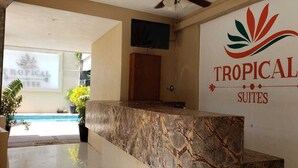 Uma área de recepção com balcão de mármore, televisão na parede e uma placa com a inscrição 'Tropical Suites'.
