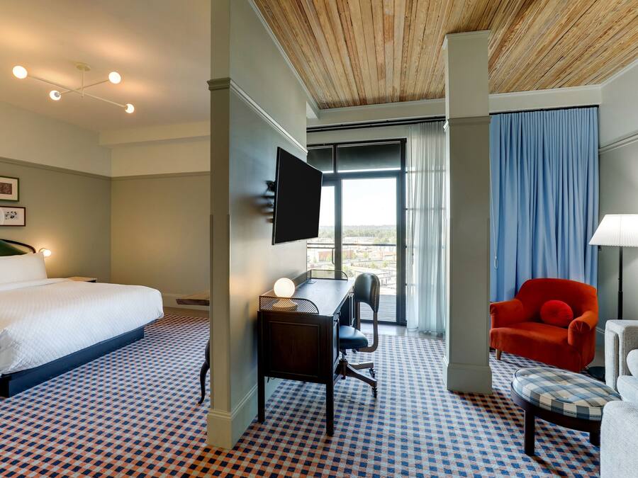 Quarto de hotel com cama, escrivaninha e área de estar. Há uma televisão na parede, uma janela com cortinas e vista para a cidade.