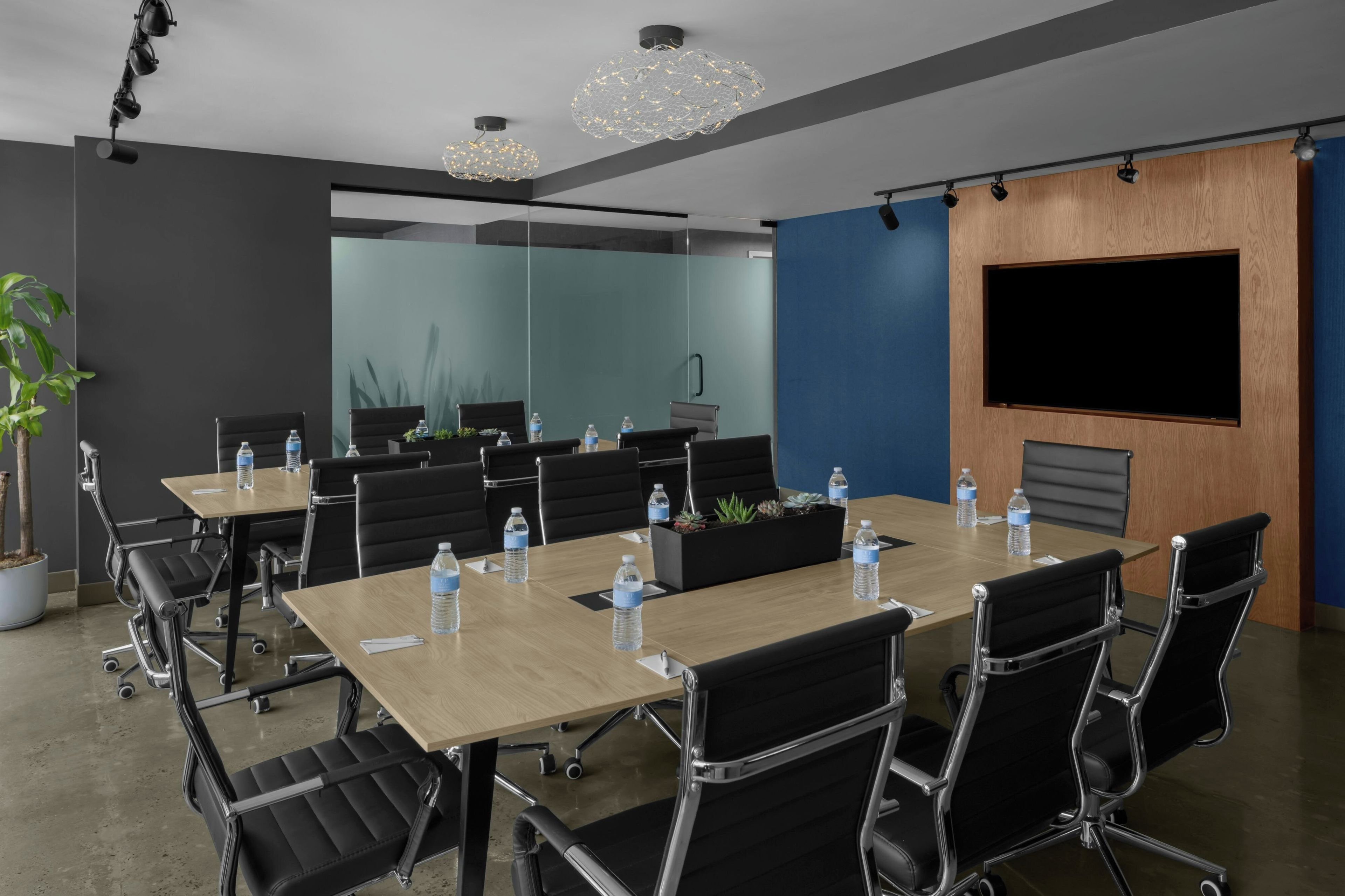Sala de conferências moderna com uma mesa comprida, cadeiras pretas, garrafas de água e um ecrã grande na parede.