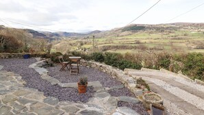 Um pátio de pedra com mesa e cadeiras, um vaso de planta e vista para um vale com casas e colinas.