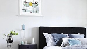 Quarto com cabeceira escura, cama com várias almofadas, mesa de cabeceira com abajur, uma pequena planta e uma obra de arte emoldurada na parede.