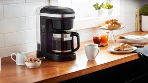Uma cozinha com cafeteira, canecas brancas, um prato de cereais, uma travessa com croissants e um copo de suco.