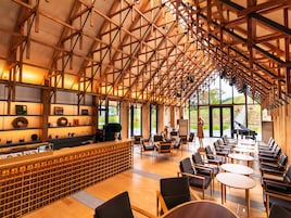 Amplo espaço interno com teto alto sustentado por vigas de madeira, área de bar com assentos e grandes janelas com vista para o exterior.