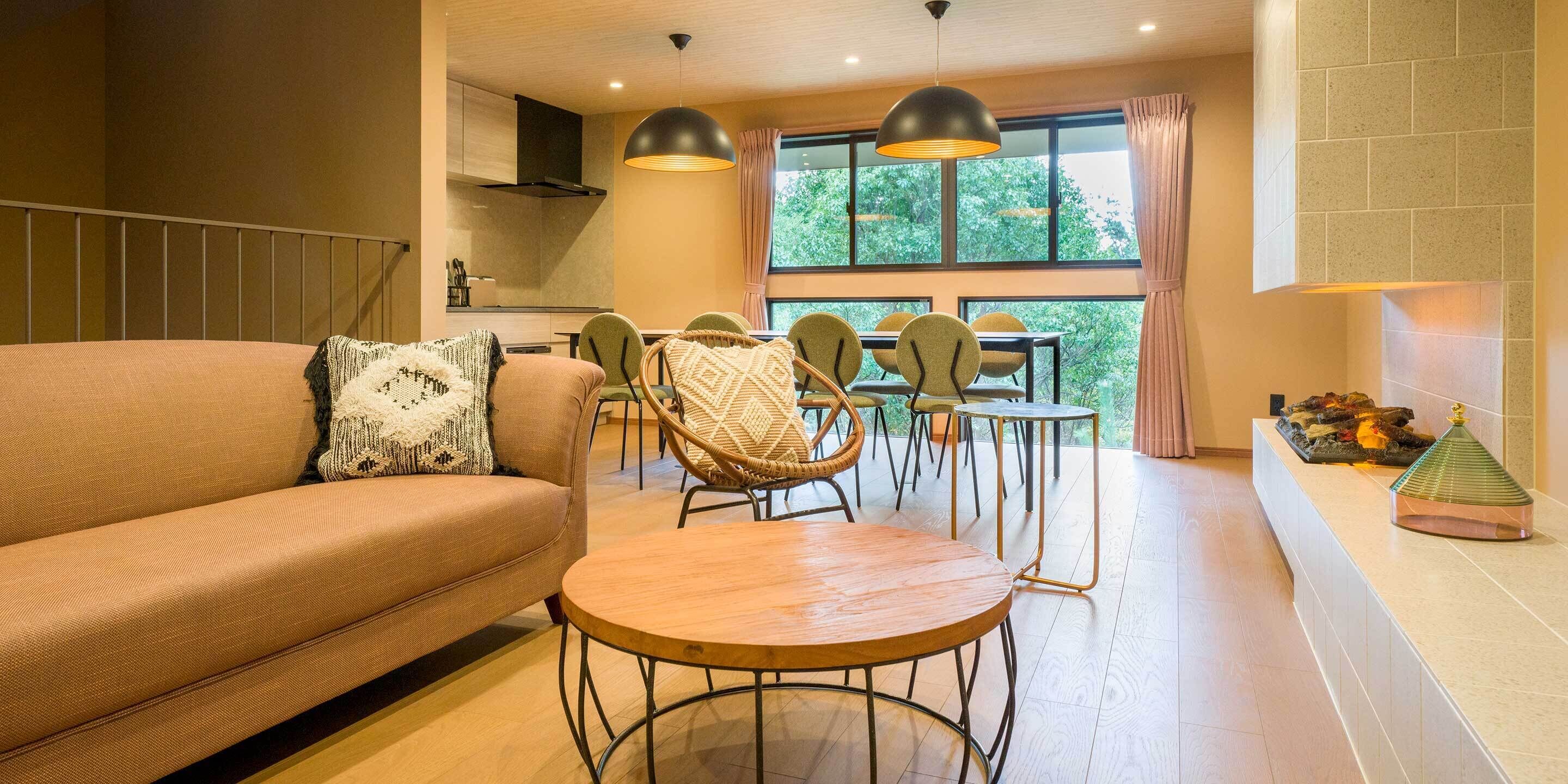 Uma sala de estar aconchegante com um sofá, uma mesa redonda de madeira e cadeiras de vime. Há uma área de cozinha com fogão e uma mesa de jantar com cadeiras. Grandes janelas oferecem vista para o verde lá fora.
