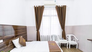 Quarto de hotel com cama, duas cadeiras brancas, uma mesinha, uma planta em vaso e cortinas.