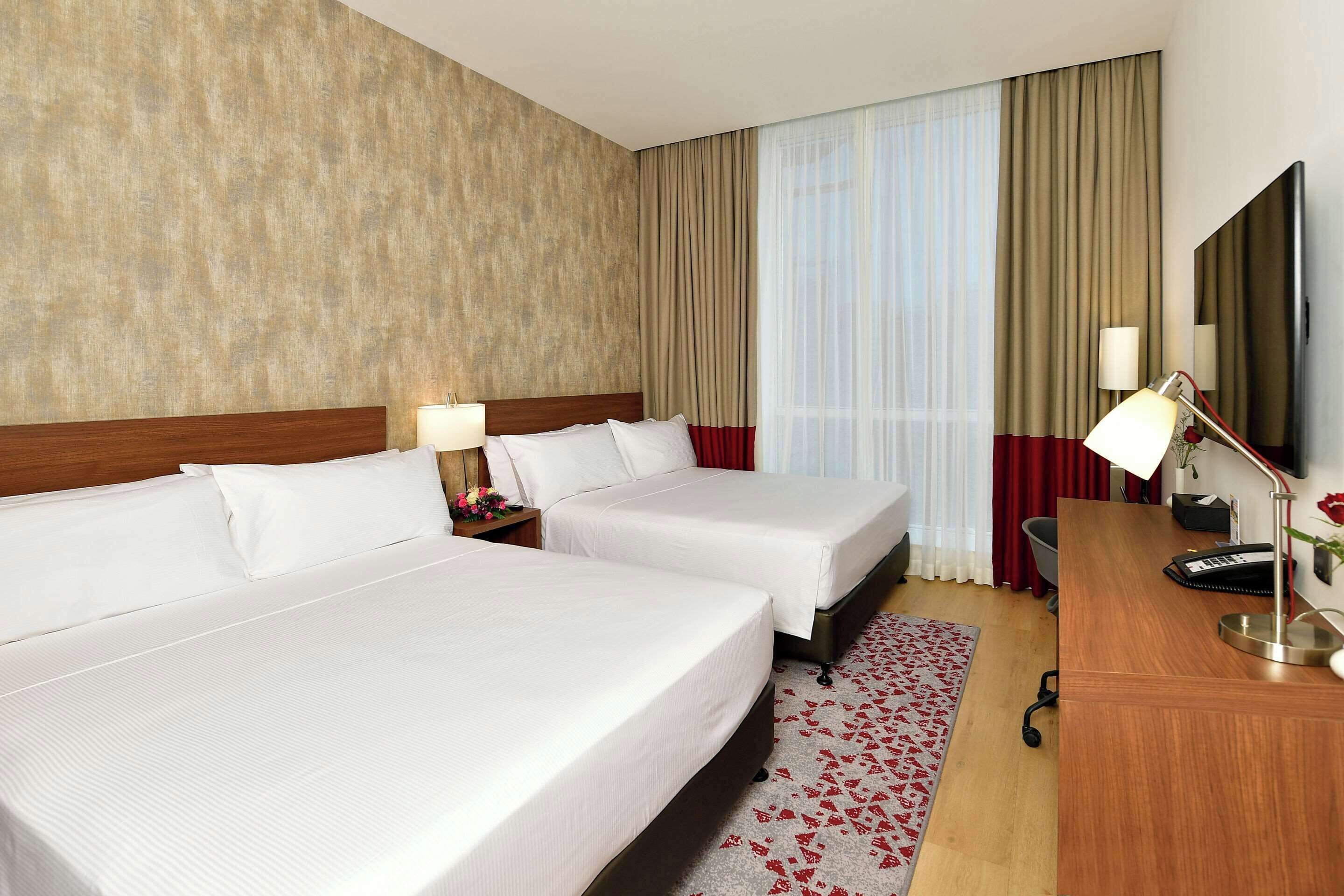 Quarto de hotel com duas camas, uma escrivaninha, uma cadeira, um abajur e um espelho grande.