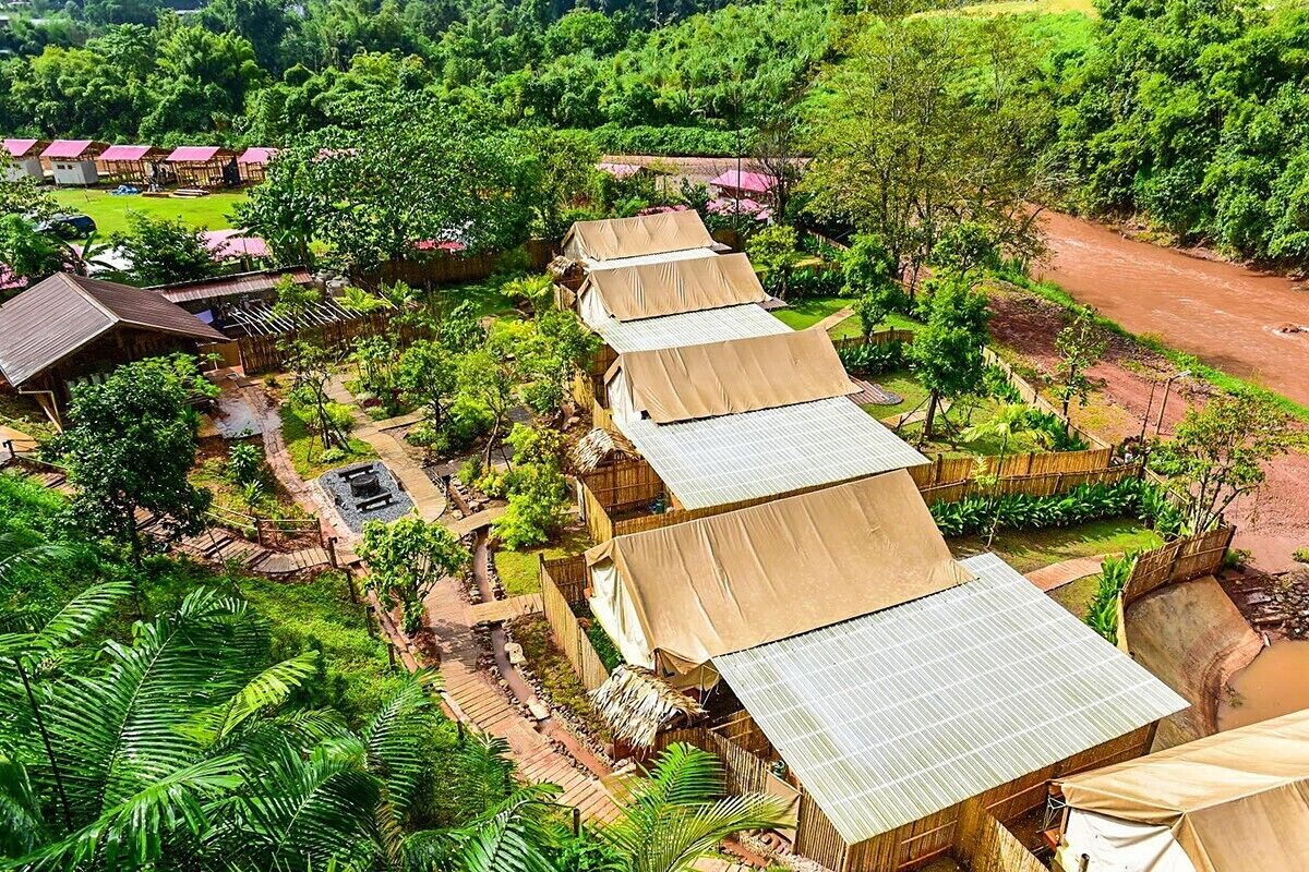 Paisagem rural com várias cabanas de telhado de palha e algumas com telhado de metal, cercada por vegetação exuberante e um rio.
