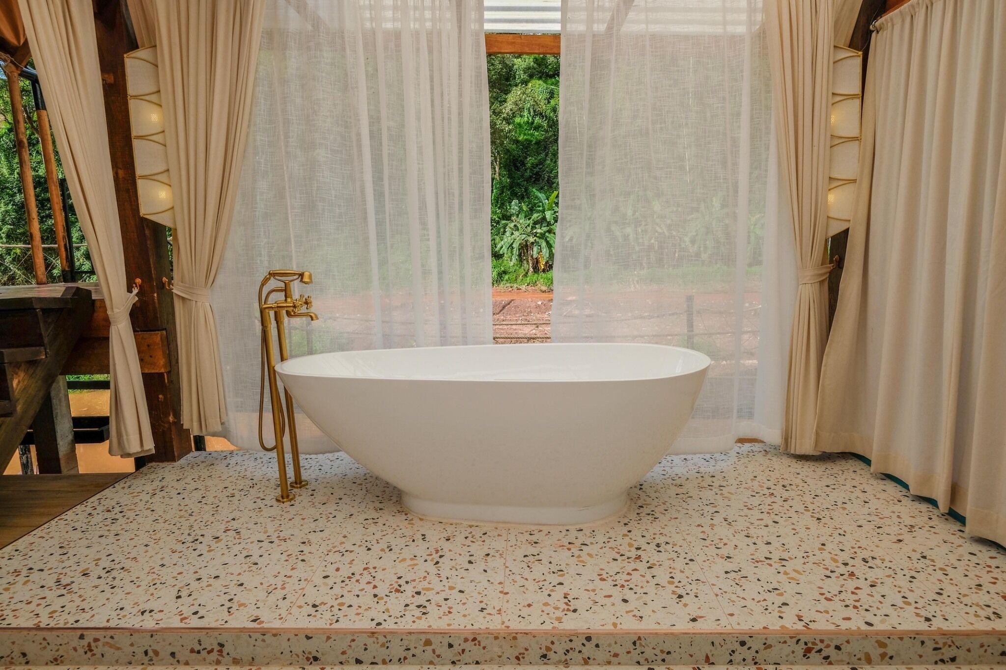 Uma banheira independente em um quarto com piso salpicado, cortinas leves e vista para vegetação lá fora.